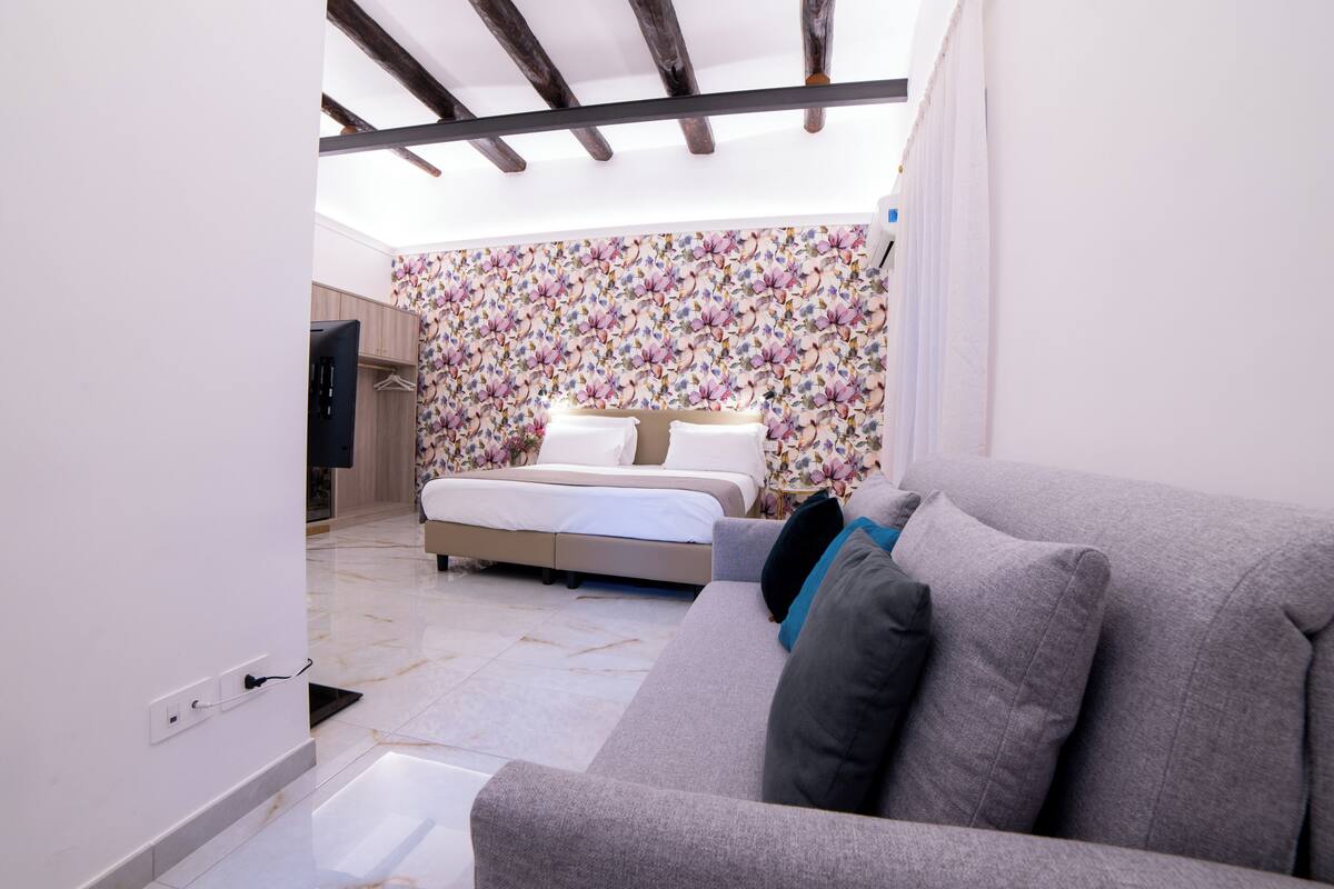 Quarto com cama, sofá cinza, parede floral e vigamento de madeira no teto.