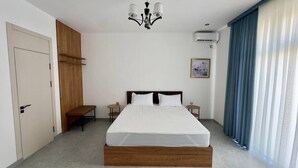 Quarto de hotel com cama, mesas de cabeceira, um banco, guarda-roupa de madeira, um candelabro, ar condicionado, um quadro emoldurado e uma janela com cortinas.