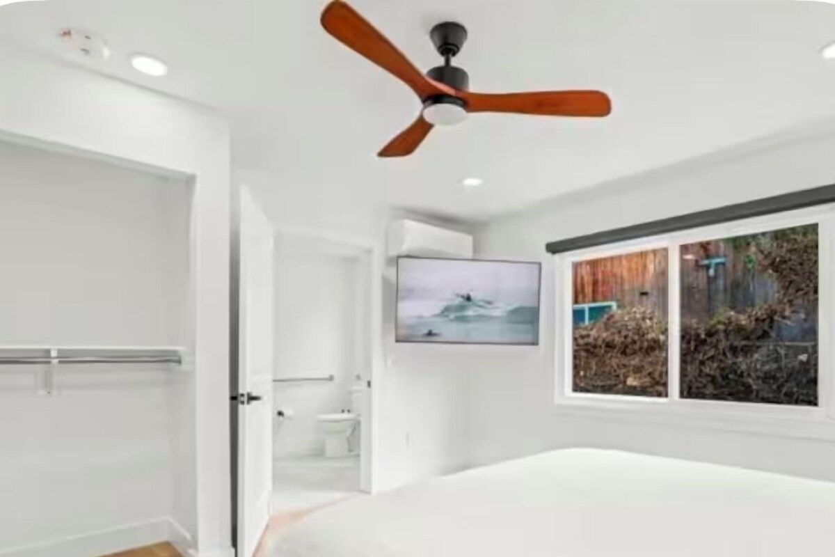 Quarto com cama, ventoinha no teto, televisão e janela com vista para árvores.