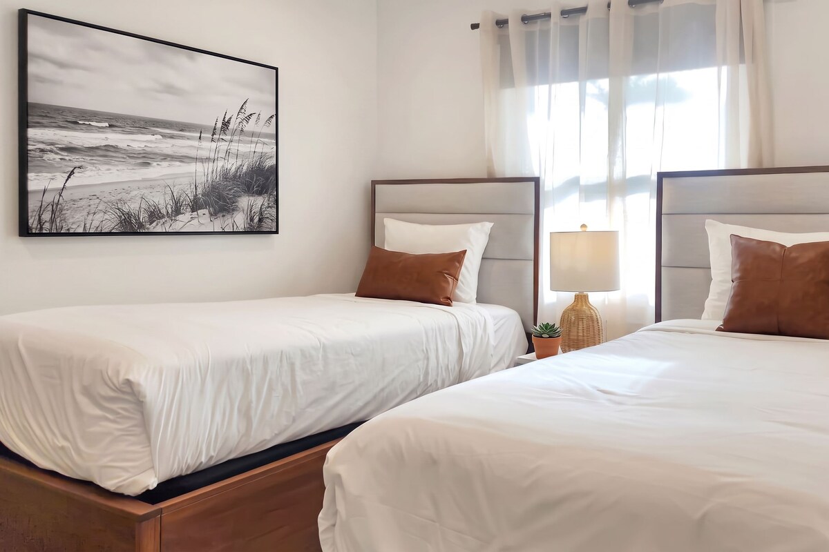Quarto de hotel com duas camas, uma mesa de cabeceira, um abajur e arte decorativa com tema praiano.
