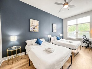 Quarto de hotel com duas camas, uma escrivaninha, uma cadeira, ventoinha no teto e obras de arte nas paredes.