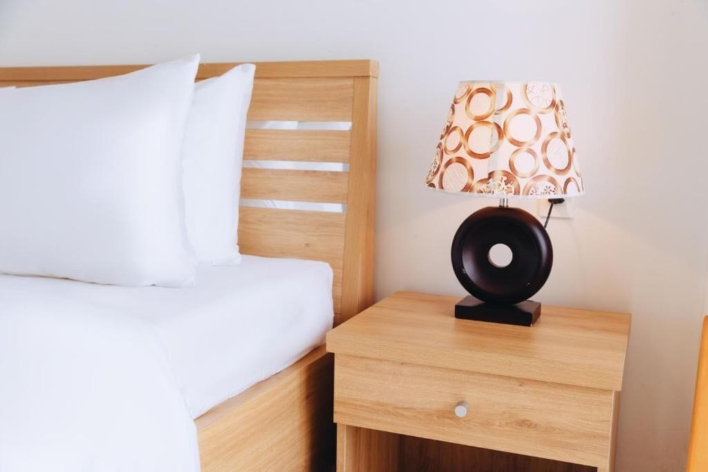 Cama com roupa de cama branca, cabeceira de madeira, mesa de cabeceira com abajur e gaveta.