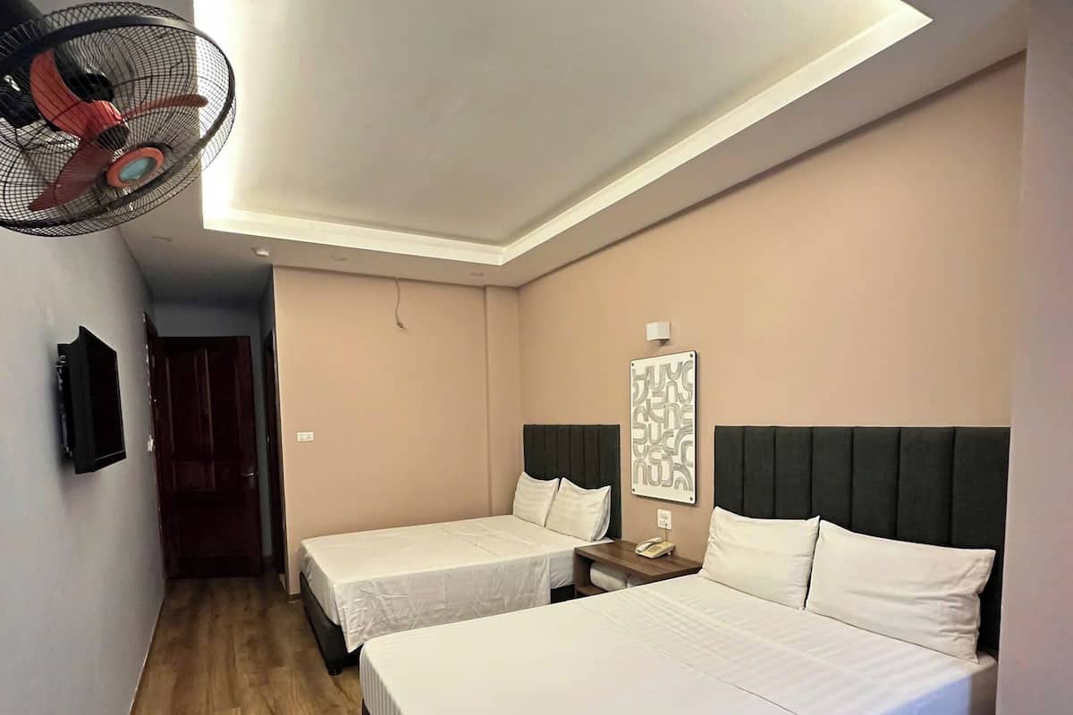 Quarto de hotel com duas camas, um ventilador de teto, uma mesa de cabeceira com abajur e uma porta que dá para outro cômodo.