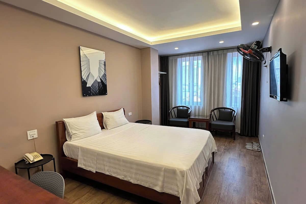 Quarto com cama, mesa de cabeceira, cadeira, televisão e janela com cortinas.