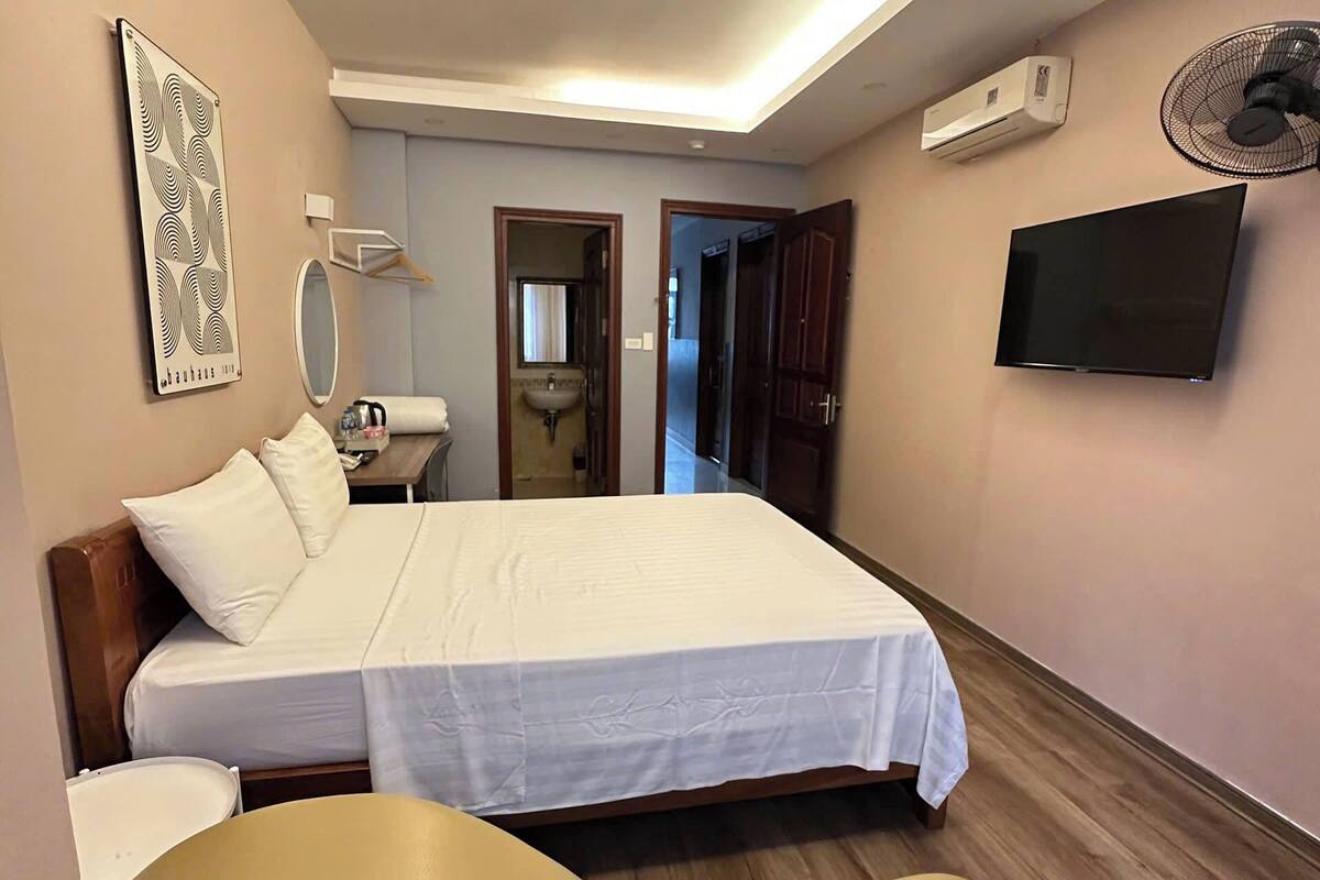 Quarto de hotel com cama, mesa de cabeceira, uma cadeira, televisão, aparelho de ar condicionado e uma porta que dá para outro cômodo.
