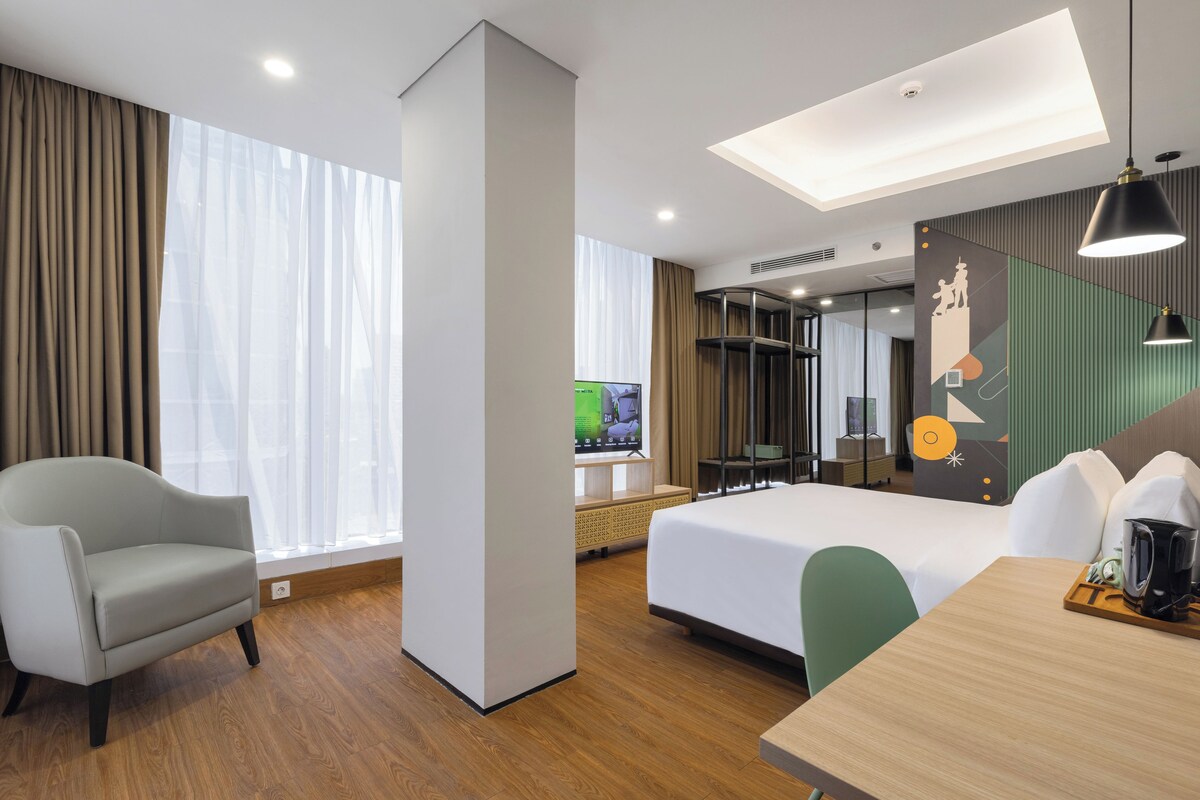 Quarto de hotel com cama, escrivaninha, cadeira, televisão e uma grande janela com cortinas.