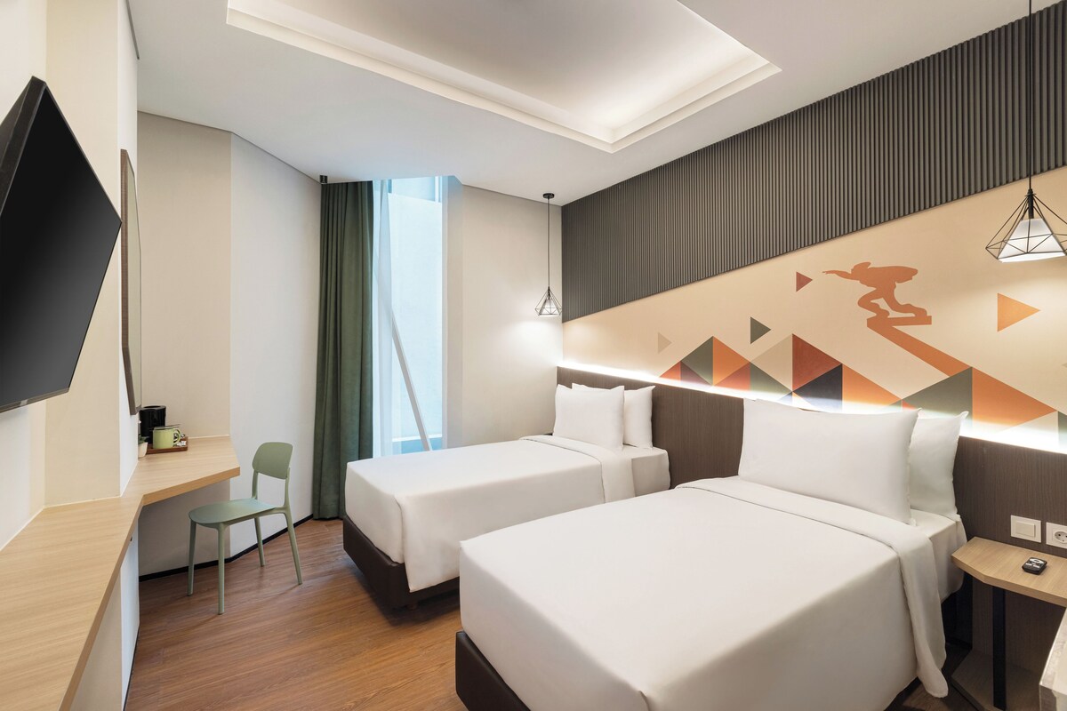 Quarto de hotel com duas camas, uma escrivaninha, uma cadeira, uma televisão e uma janela com cortinas.