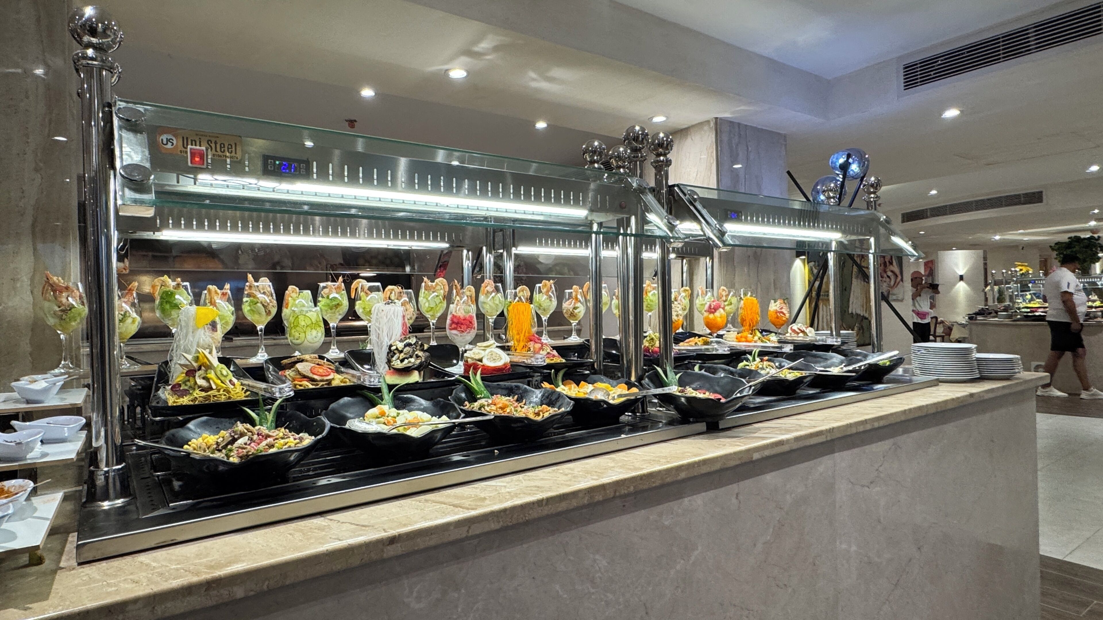 Buffet com diversos alimentos e um relógio digital mostrando a hora.