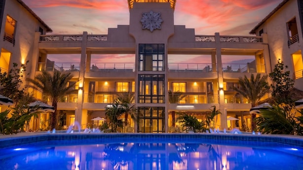 Um hotel de vários andares com entrada central, piscina e palmeiras.