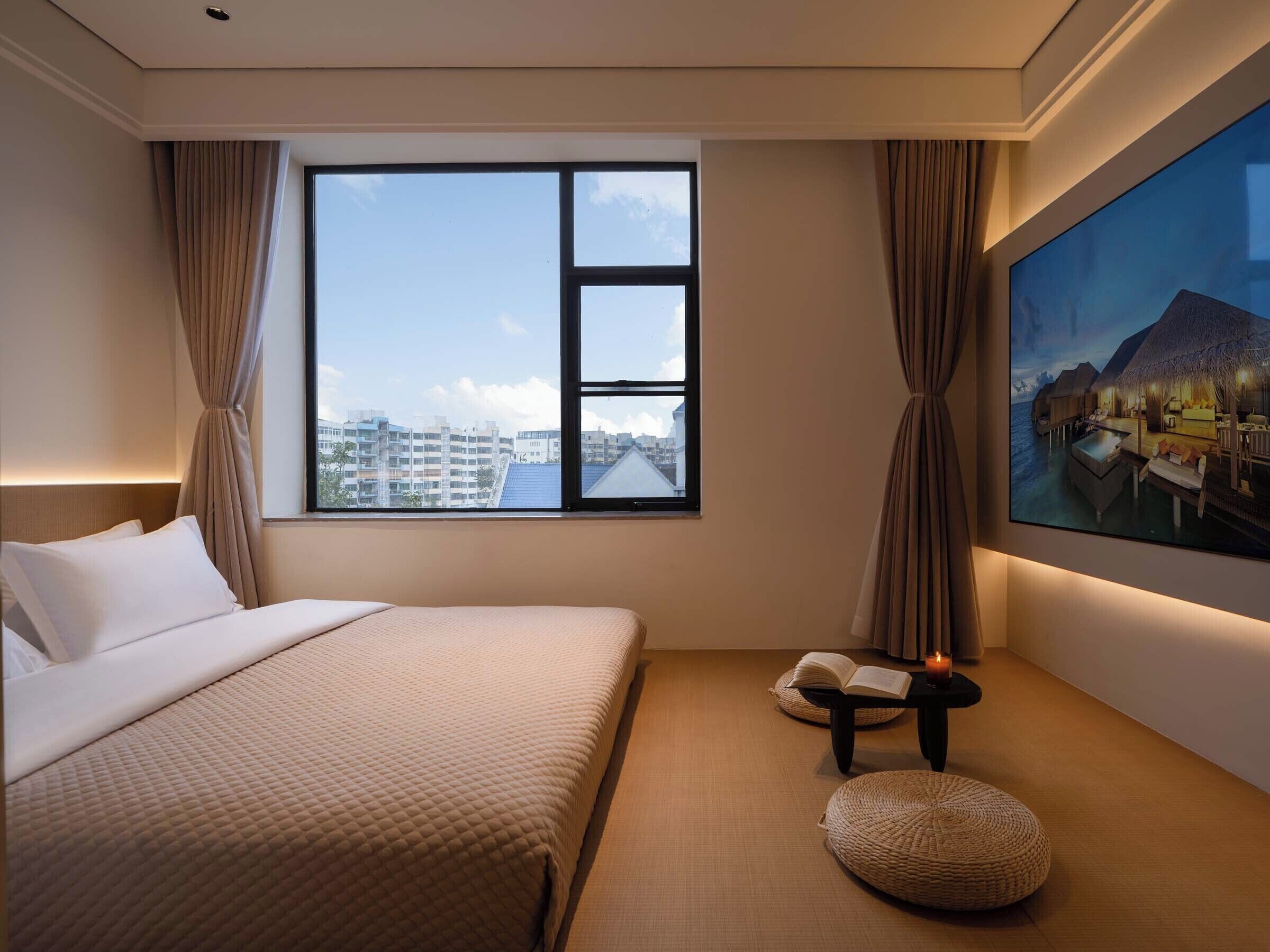 Quarto de hotel com uma cama grande, janela com cortinas, uma mesinha com um livro e uma televisão presa à parede.