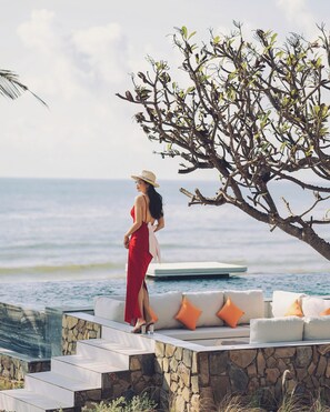 Mulher de vestido e chapéu vermelho parada num terraço de pedra com vista para um corpo de água.