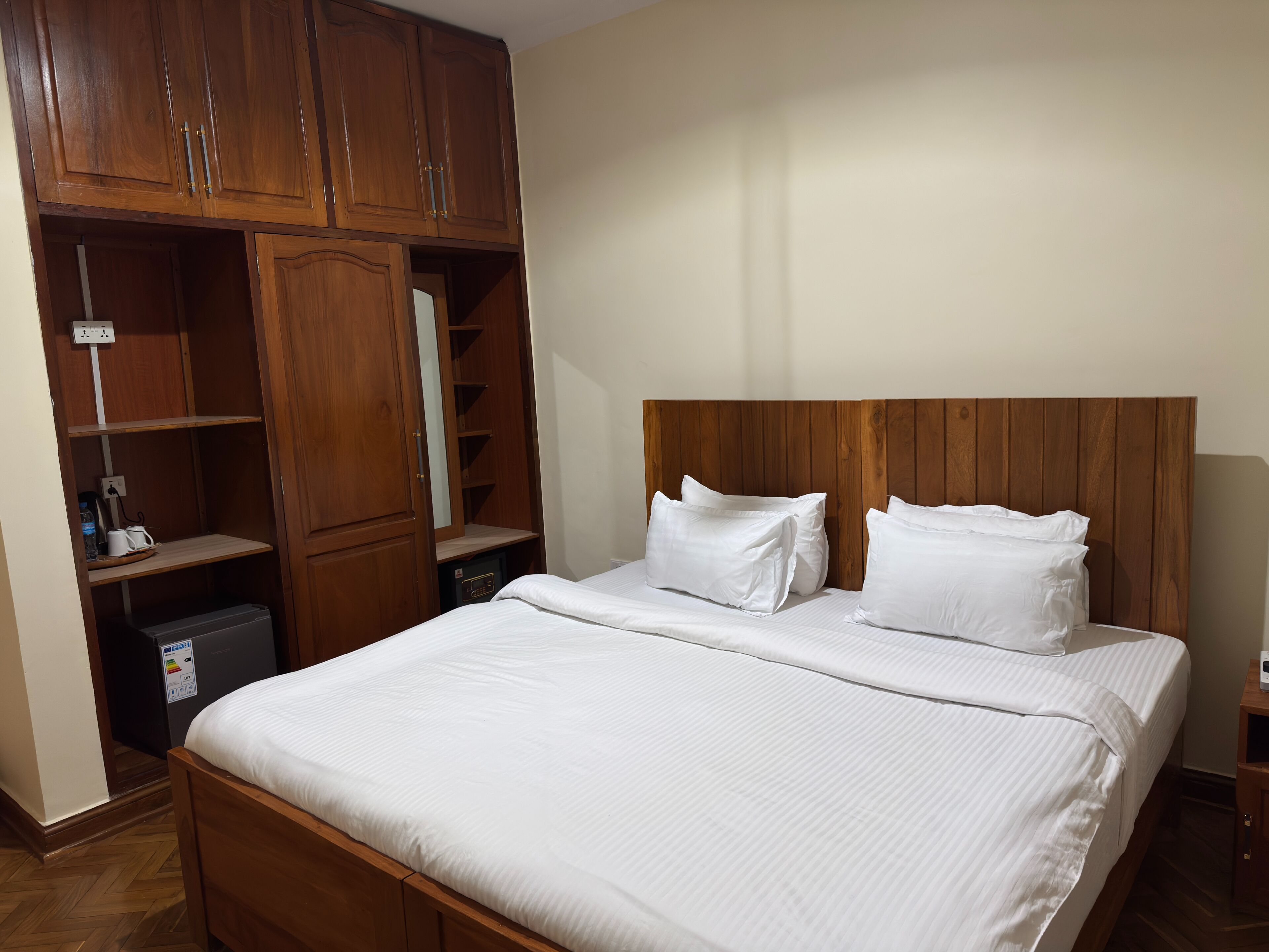 Quarto com cabeceira de madeira, duas almofadas brancas, cama com colcha branca, guarda-roupa de madeira e uma pequena mesa de cabeceira.
