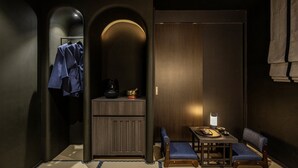 Quarto pequeno com guarda-roupa, uma mesinha com abajur, uma cadeira e um cabideiro com roupas.