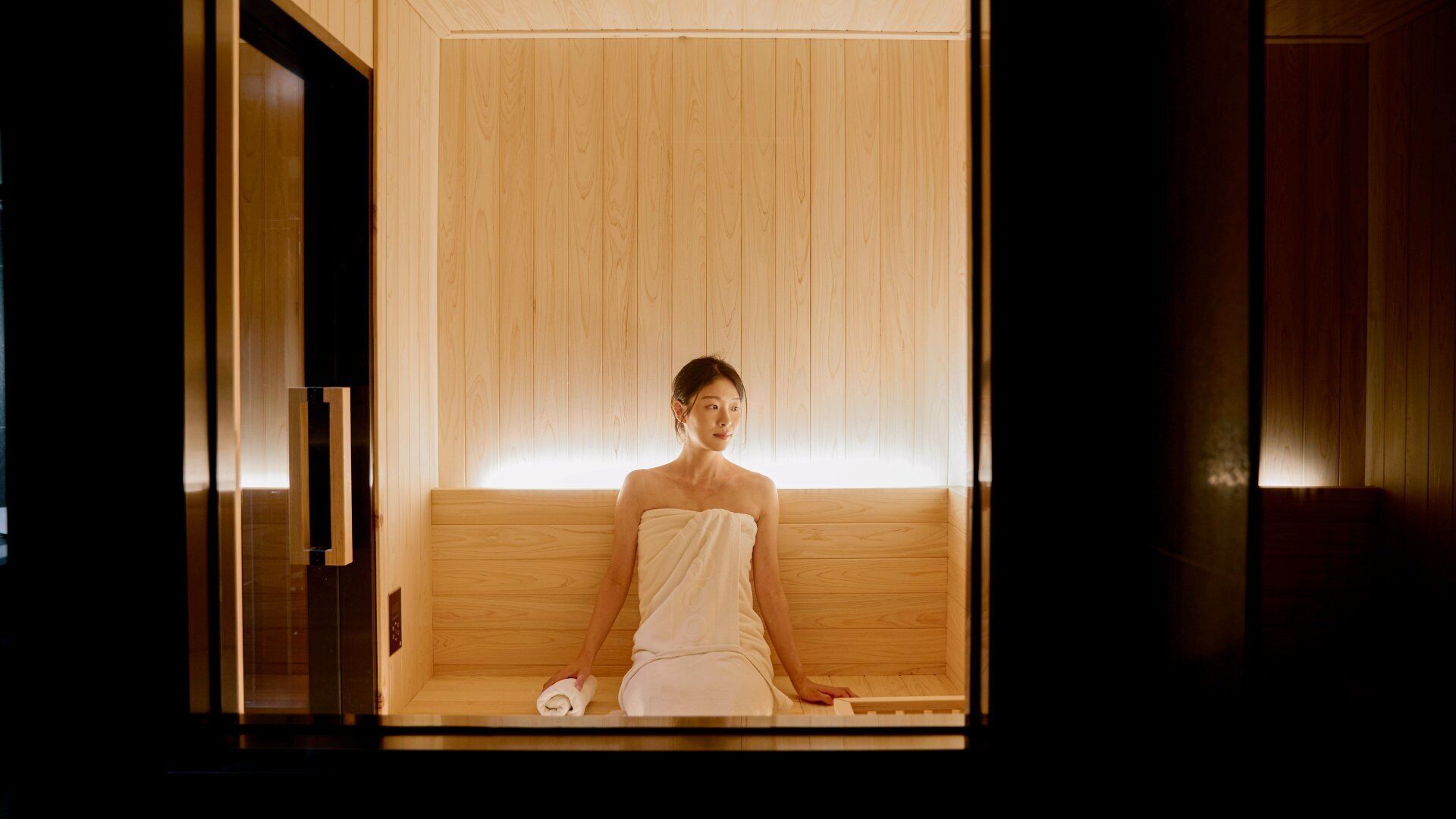 Uma pessoa sentada em uma sauna de madeira com uma toalha enrolada ao corpo.