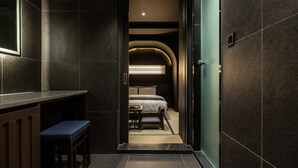 Um corredor estreito que dá para um quarto com cama, mesa de cabeceira e um banco.