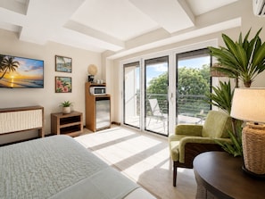 Quarto de hotel com cama, escrivaninha, mini-geladeira, micro-ondas, televisão e porta de vidro corrediça que dá para uma área externa com uma cadeira e uma mesa.