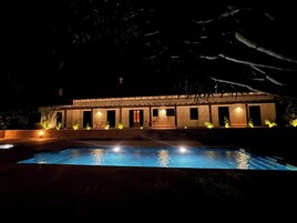 Vista noturna de uma casa com piscina, apresentando um espaço coberto de varanda e várias janelas.