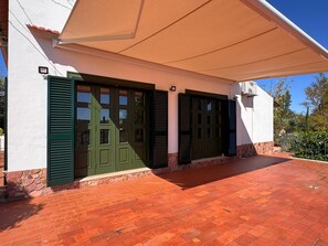Um edifício com varanda coberta, persianas verdes e piso de tijolos.