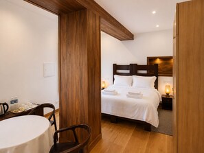 Quarto de hotel com cabeceira de madeira, cama com roupa de cama branca, mesa redonda com uma cadeira e abajur.
