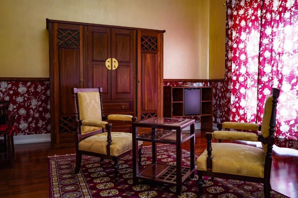 Quarto com um guarda-roupa de madeira, duas poltronas amarelas, uma mesa de madeira e uma cortina floral vermelha.