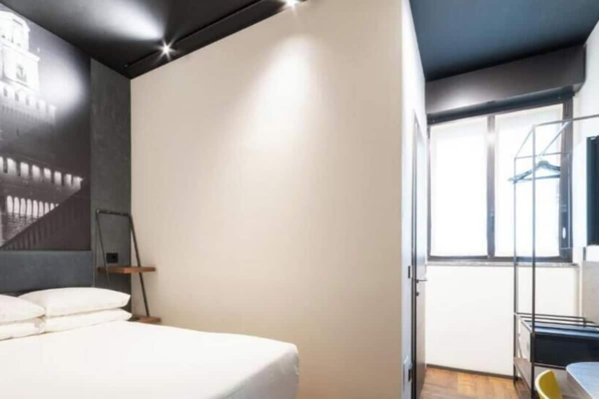 Quarto de hotel com cama, escrivaninha, cadeira, luminária e uma grande janela.