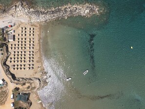 Vista aérea de uma praia com fileiras de espreguiçadeiras, costa rochosa e barcos na água.
