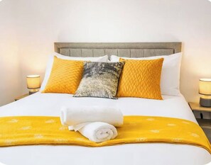 Uma cama com cabeceira, duas luminárias de apoio e uma toalha enrolada sobre um cobertor amarelo.