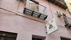 Um edifício com fachada rosa, uma placa com 'B&B' e informações de contato, uma varanda com grade de ferro forjado e um aparelho de ar condicionado.