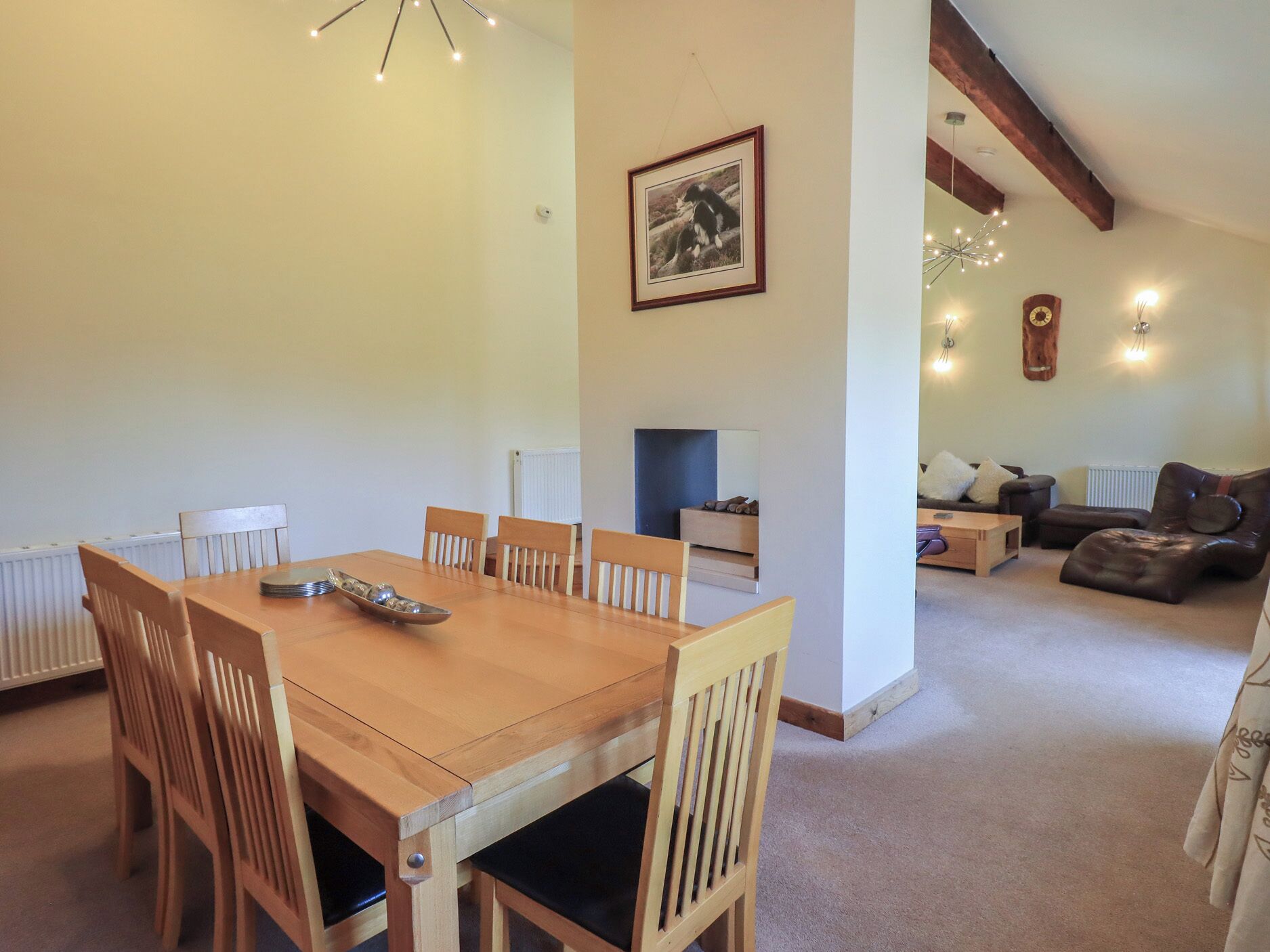 Uma sala de jantar com uma mesa e cadeiras de madeira, uma lareira, e uma sala de estar com um sofá e um relógio na parede.