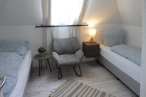 Quarto pequeno e bem arrumado, com uma única cadeira, mesa de cabeceira com luminária e duas camas.