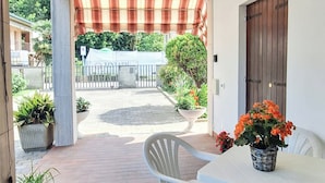 Um alpendre coberto com mesa, cadeiras, plantas em vasos e vista para a rua.