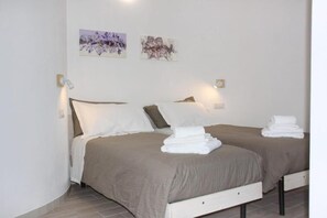 Uma cama de casal com colcha bege, dois travesseiros e uma toalha dobrada em cada cama. Há duas luminárias fixadas na parede e duas pinturas abstratas.