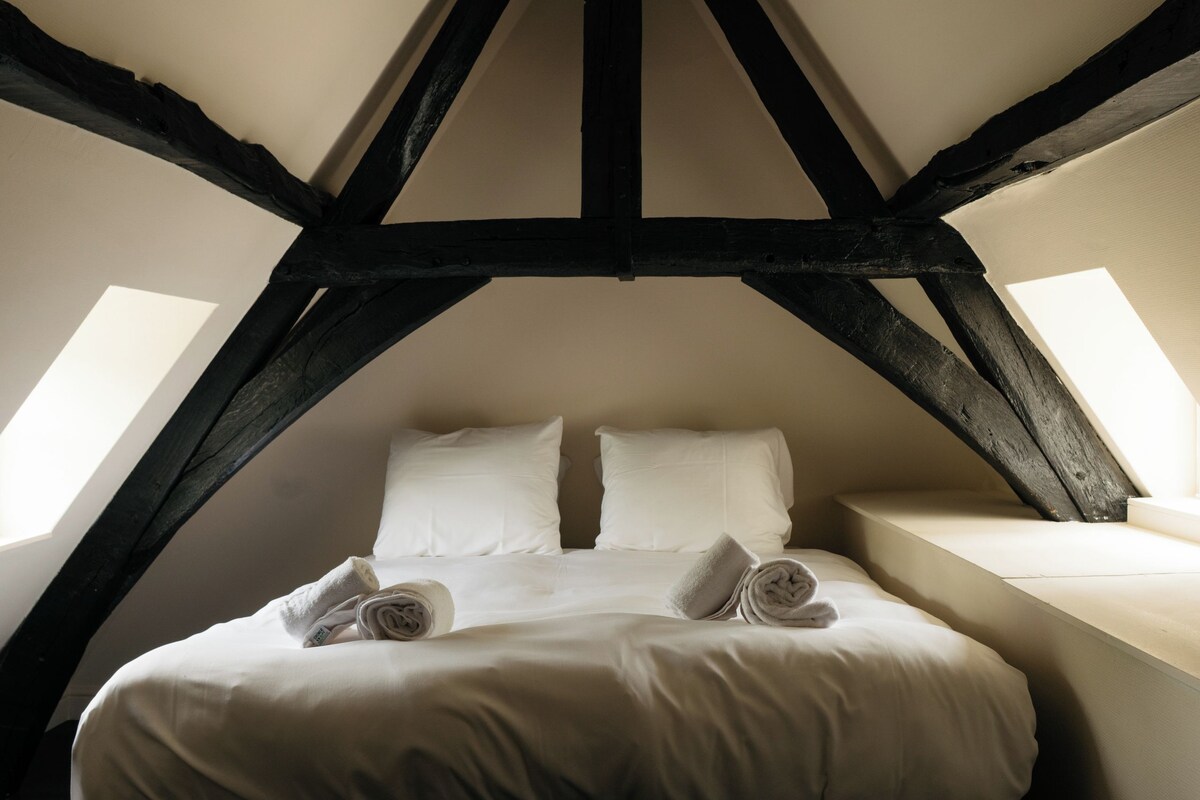 Quarto com teto inclinado, cama com roupa de cama branca e duas claraboias.