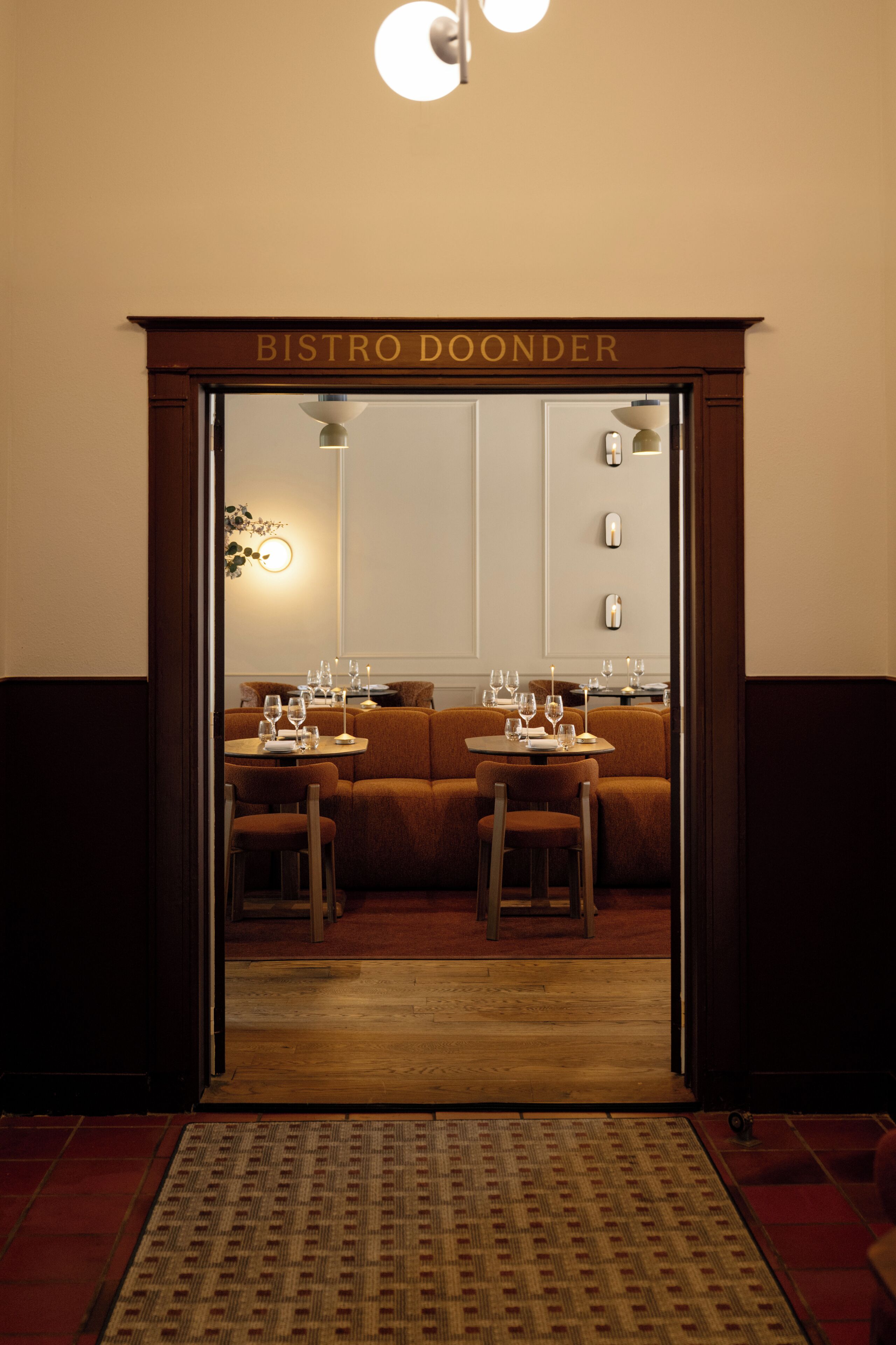 Entrada de um restaurante com uma placa que diz 'BISTRO DOONDER'. No interior, mesas montadas com copos e talheres, e um sofá é visível ao fundo.