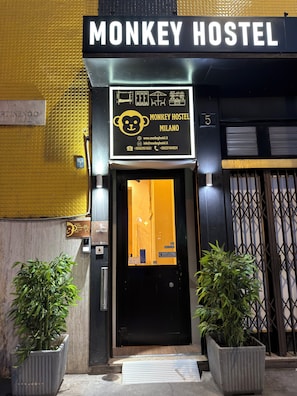 Exterior do Monkey Hostel em Milão, com uma placa de destaque com o logotipo de um macaco, o número 5 e informações de contato.