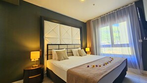 Quarto de hotel com uma cama grande, uma mesa de cabeceira com abajur, uma janela com cortinas e vista para o verde lá fora.