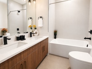 Uma casa de banho moderna com lavatório branco, torneiras pretas, toucador de madeira, sanita branca e banheira.