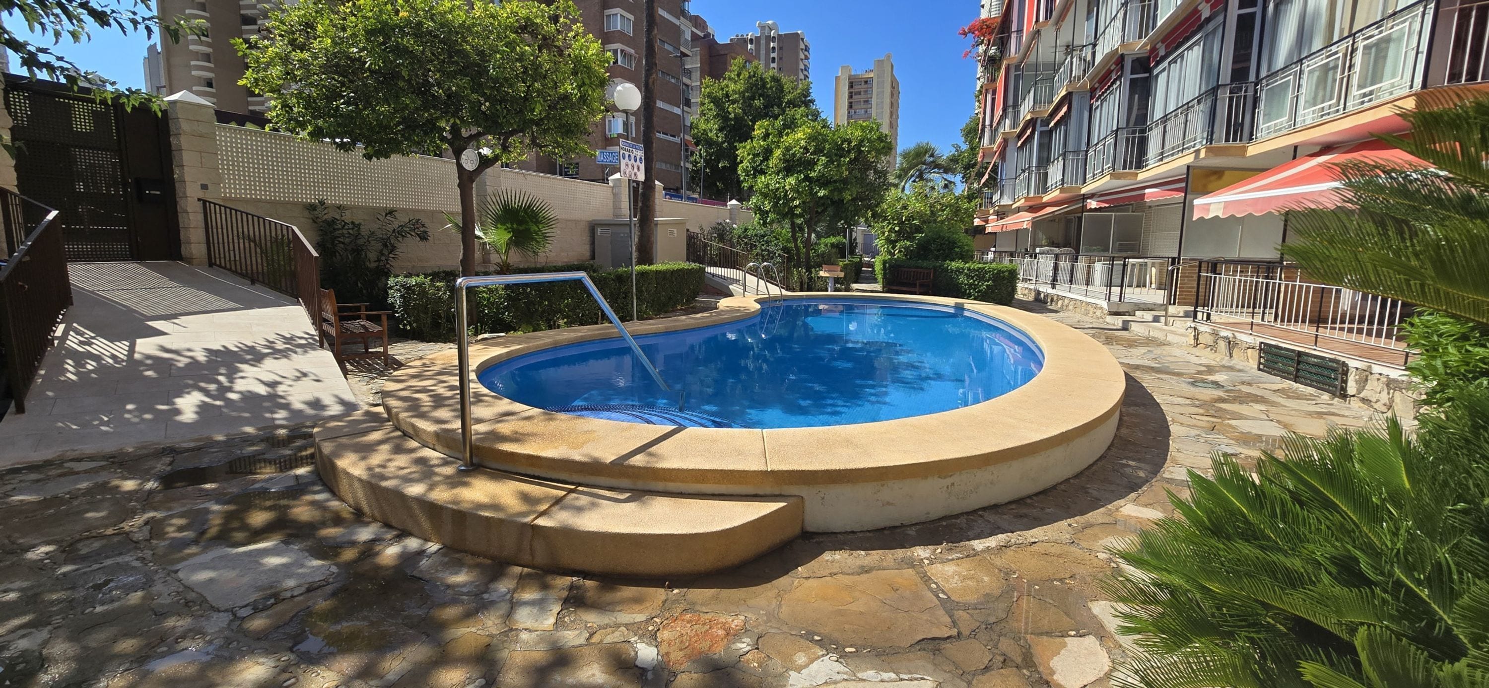Uma piscina com borda de pedra, rodeada por um terraço de pedra, árvores e edifícios altos ao fundo.
