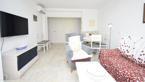Uma sala de estar moderna com uma TV de tela plana, um sofá, uma mesa de jantar com cadeiras e uma poltrona cinza.