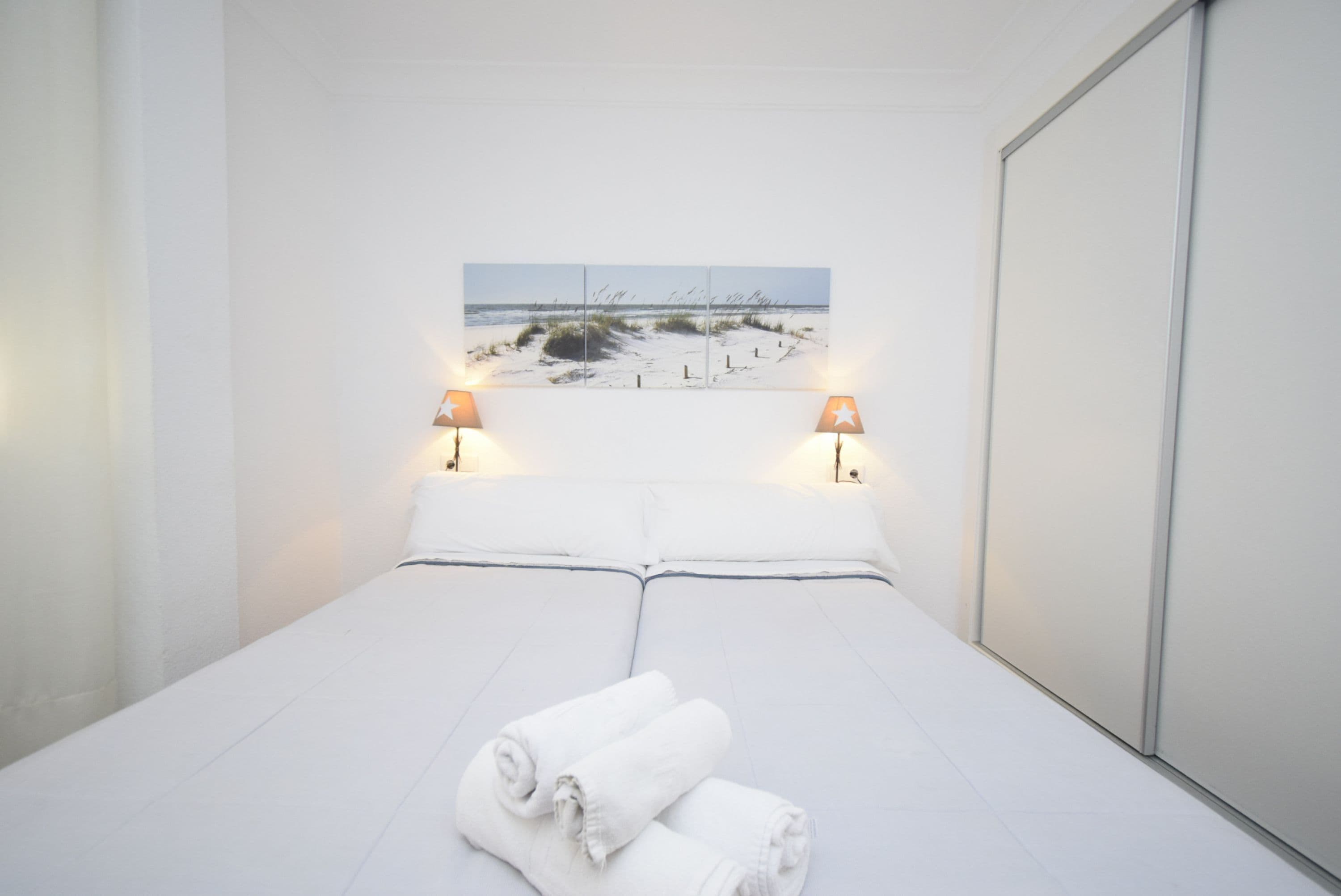 Quarto de hotel com uma cama, duas luminárias de cabeceira, vista para a praia e uma toalha dobrada sobre a cama.