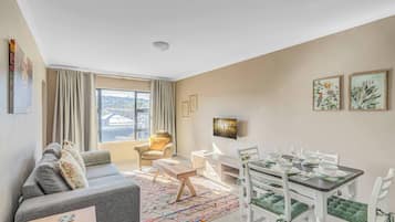 Uma sala de estar moderna com sofá, mesa de jantar posta para quatro pessoas, televisão na parede e uma janela grande com cortinas.