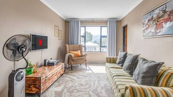Uma sala de estar com televisão, ventoinha, sofá, cadeira, janela com cortinas e um quadro na parede.