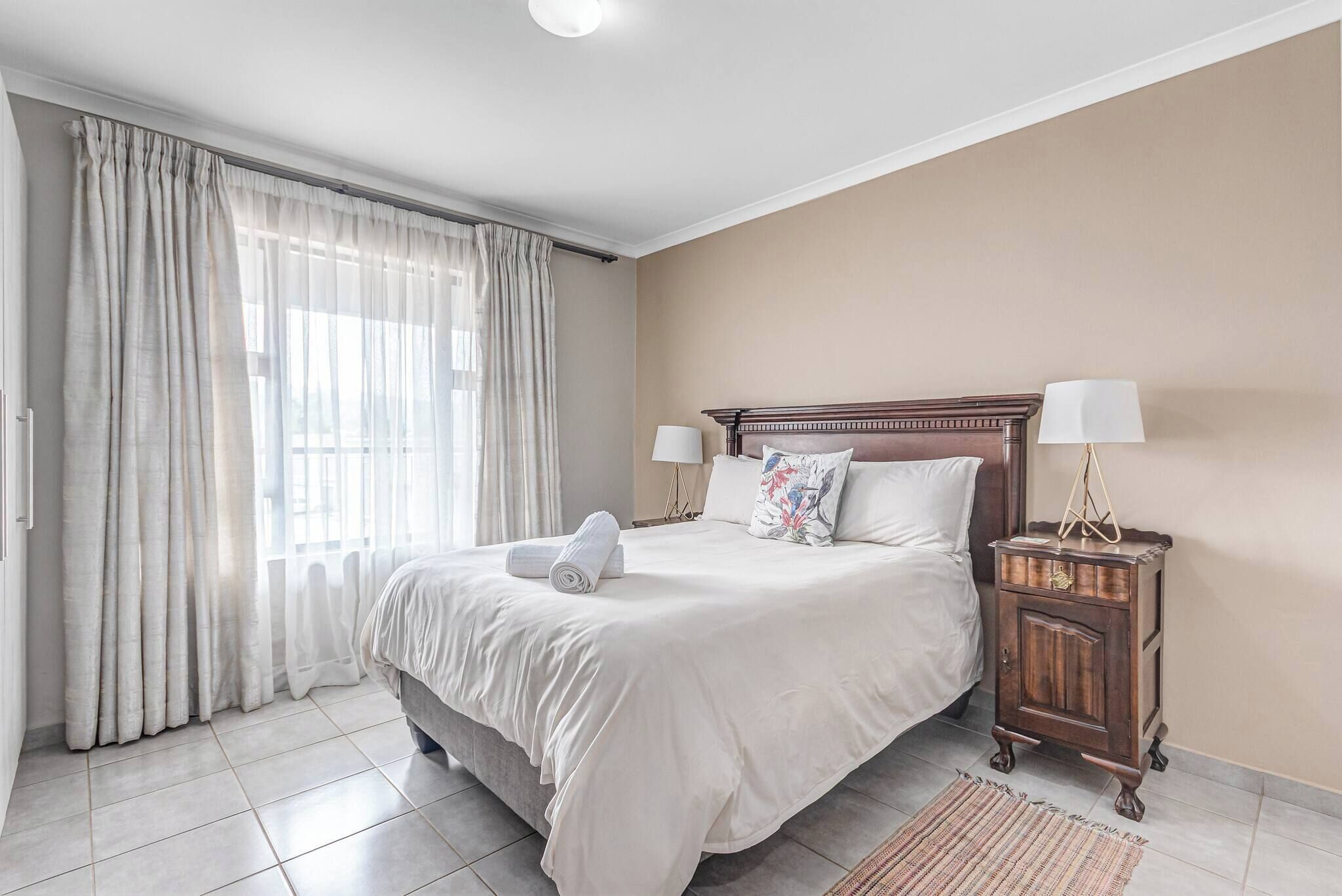 Quarto de hotel com uma cama grande, uma mesa de cabeceira com luminárias e vista para o exterior através de cortinas leves.