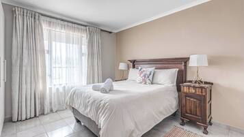 Quarto de hotel com uma cama grande, uma mesa de cabeceira com luminárias e vista para o exterior através de cortinas leves.