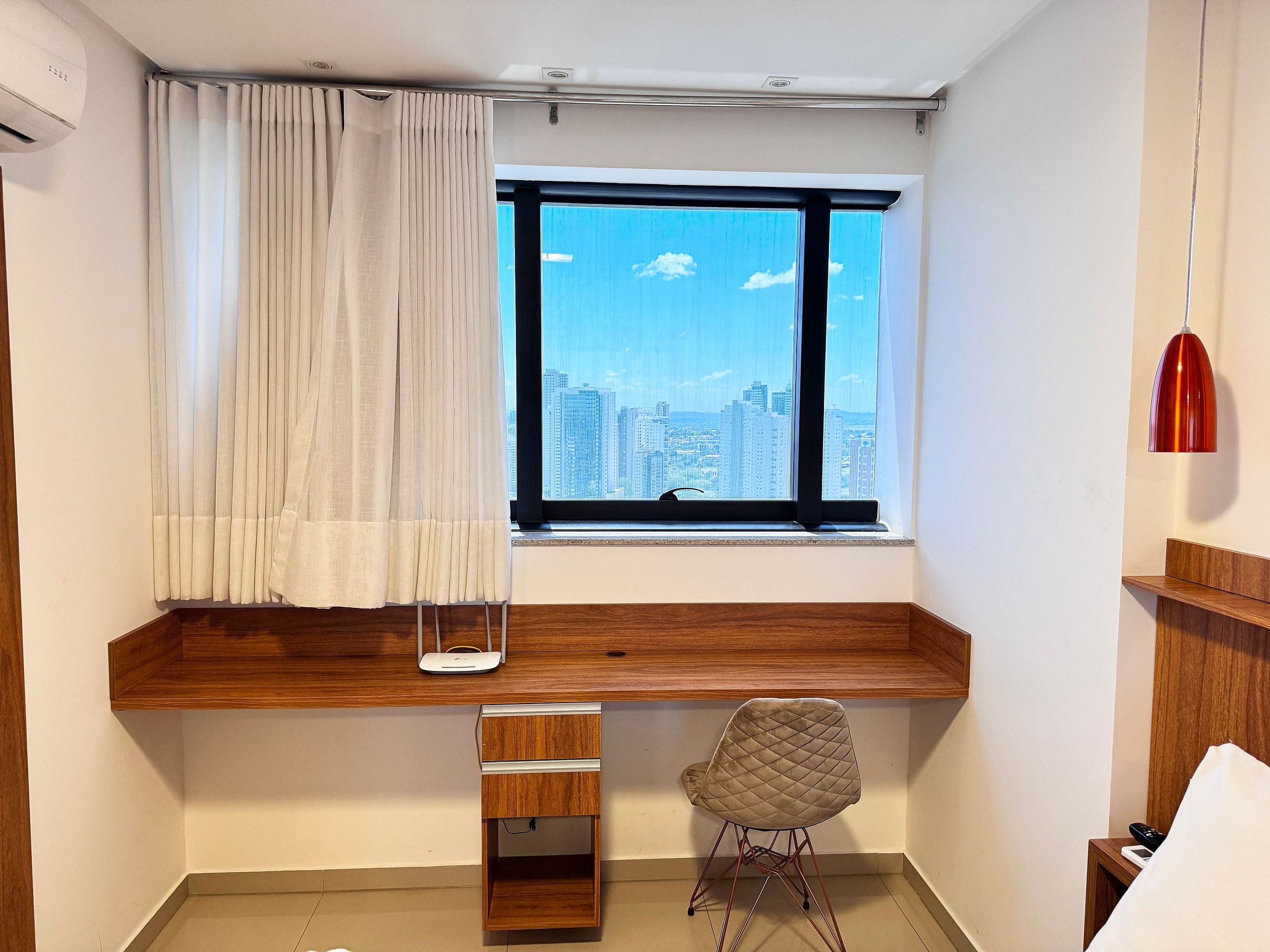 Quarto com vista para a cidade, uma escrivaninha com cadeira, uma luminária pendente vermelha e um ar condicionado branco.