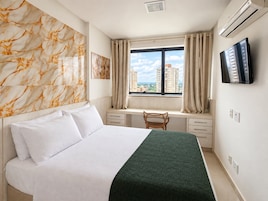 Quarto de hotel com cama, escrivaninha, cadeira, televisão e janela com vista para a cidade.