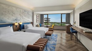 Quarto de hotel com duas camas, uma escrivaninha, televisão e uma janela grande com vista para o exterior.