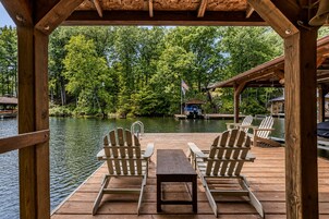 Um deck de madeira com cadeiras Adirondack e uma mesa, com vista para um lago com barcos atracados e uma cabana pequena.