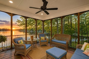 Um deck coberto ao ar livre com móveis de vime, um ventilador de teto e vista para um lago ao pôr do sol.