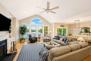 Uma sala de estar ampla com lareira, uma grande janela com vista para um lago, um ventilador de teto e um espaço de jantar também com vista para o lago.